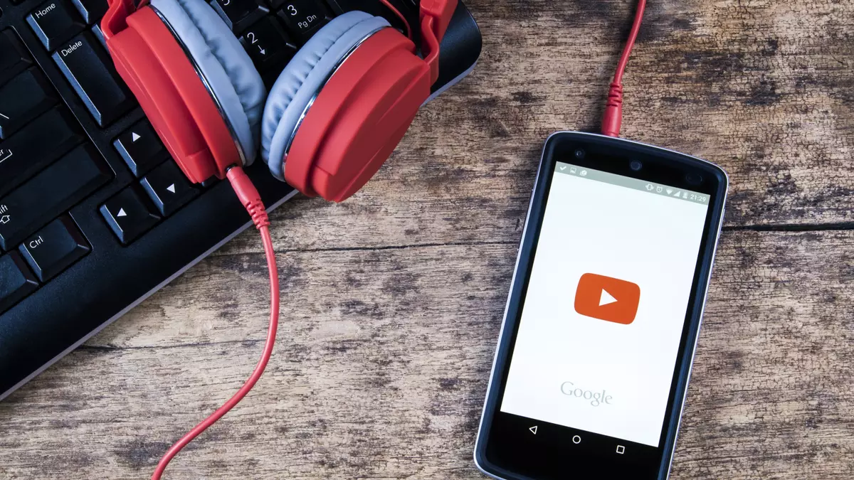 Smartphone kun ŝarĝo de YouTube Music app sur ekrano kuŝanta sur skribotablo kun aŭdiloj