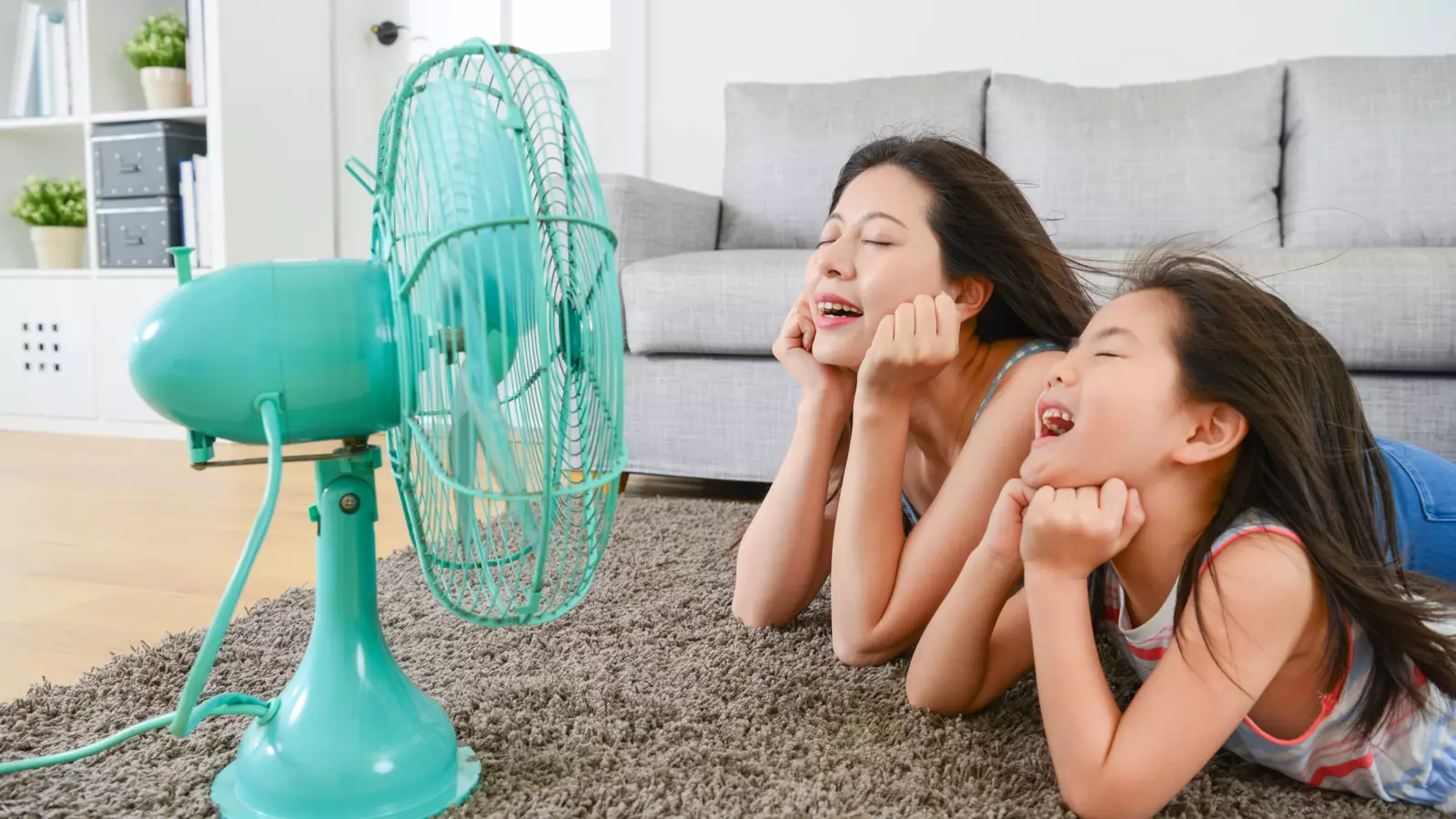 пријатна мајка са малом ћерком који леже на поду дневне собе и гледају на електрични вентилатор како заједно уживају у хладном ветру током лета