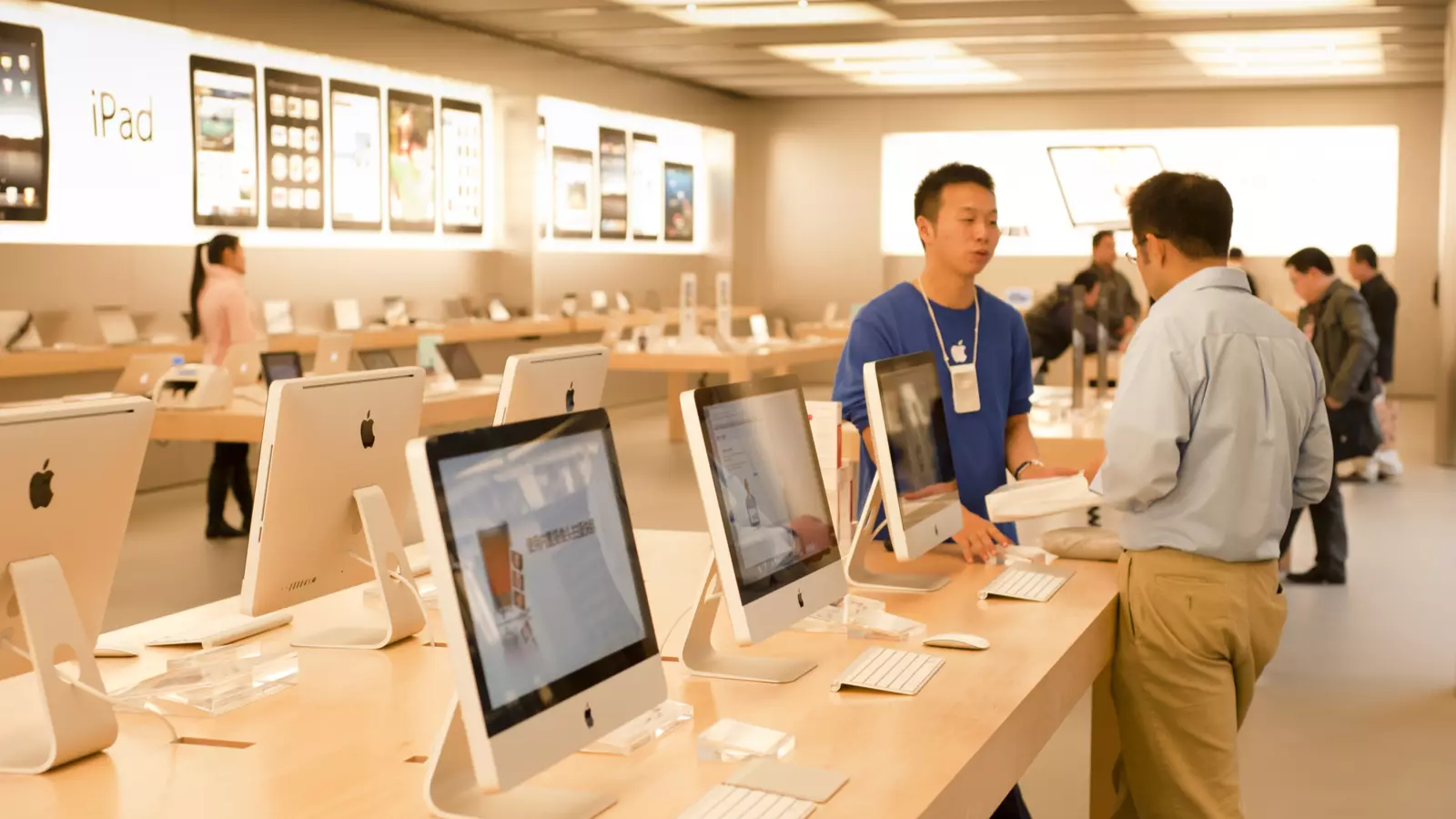 Apple စတိုးအတွင်းပိုင်းဆိုင်ရှိ iMacs ဘေးရှိ သုံးစွဲသူတစ်ဦးနှင့် Apple အရောင်းလက်ထောက် လူငယ်တစ်ဦး စကားပြောနေပါသည်။