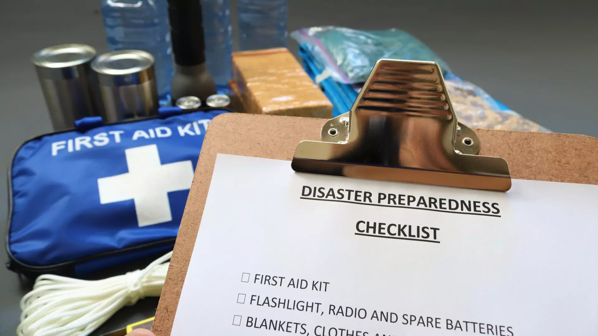 剪貼板上的備災清單，背景是救災物品。這些物品包括急救箱、手電筒、罐頭食品、水、電池和住所。
