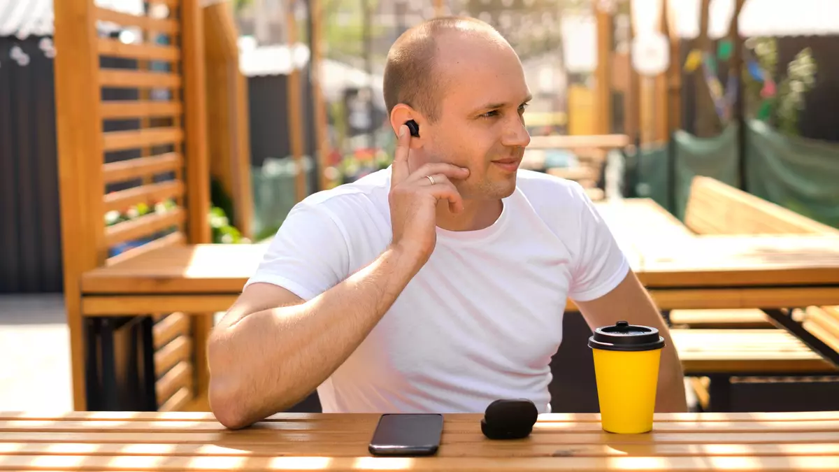 Los hombres incluyen el dedo en los auriculares inalámbricos dentro de la oreja. Experiencia agradable Escuchar música con auriculares en un café en la terraza de la ciudad en una fresca mañana de verano.