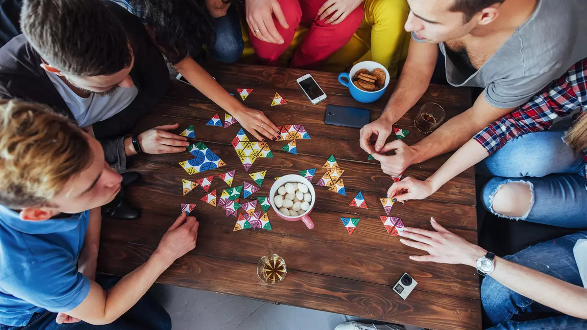 Vista superior de la foto creativa d'amics asseguts a la taula de fusta jugant a un joc de taula amb aperitius i begudes