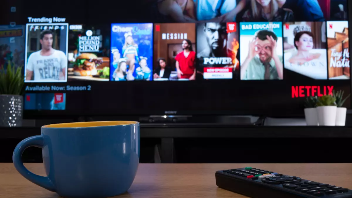 Uma caneca de café e controle remoto de TV na frente de uma TV mostrando as opções do Netflix