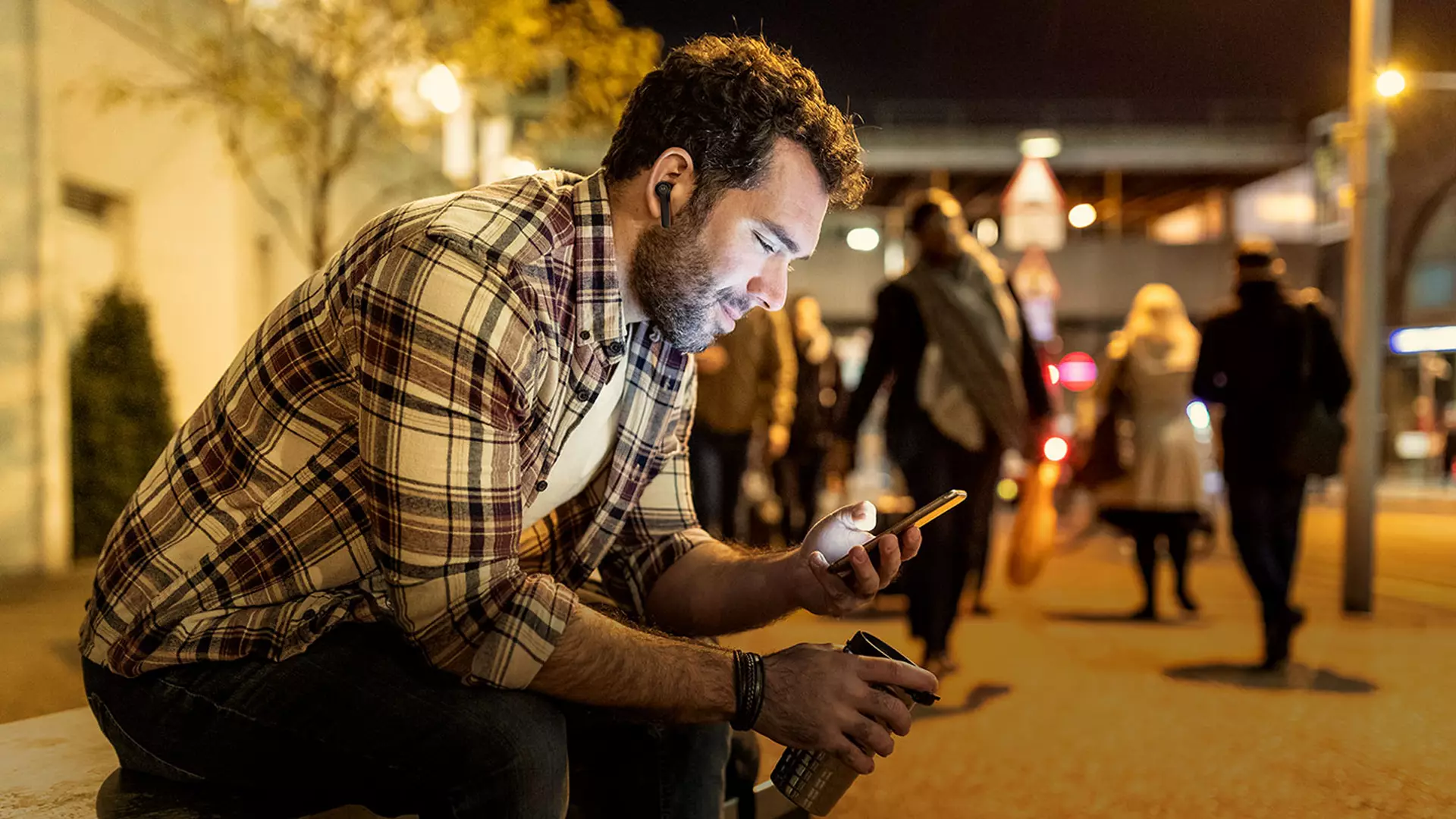 Osoba, ktorá sedí v noci vonku v meste a nosí Moto Buds, pozerá sa na svoj telefón a usmieva sa.