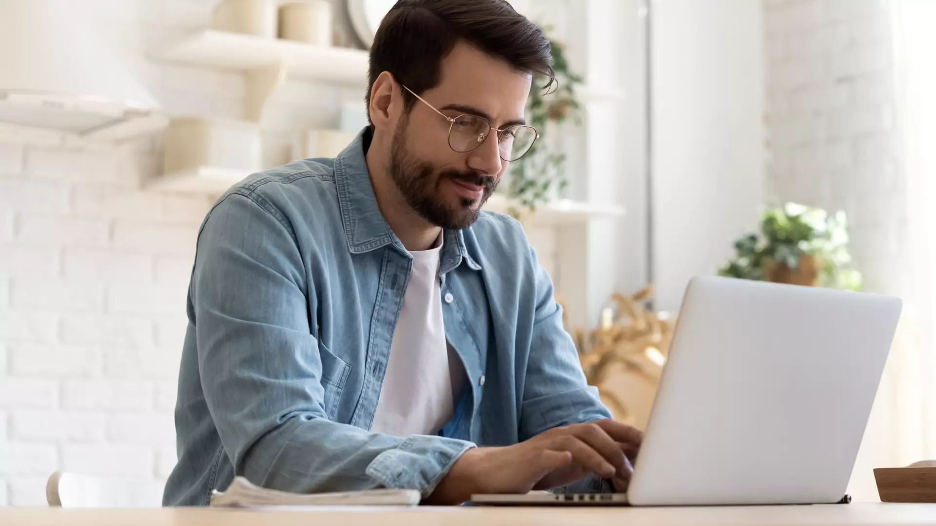 Fokussierter junger Mensch, der eine Brille mit Laptop trägt, auf der Tastatur tippt, E-Mails oder Nachrichten schreibt, chattet, einkauft, erfolgreicher Freiberufler, der online am Computer arbeitet und in einer modernen Küche sitzt