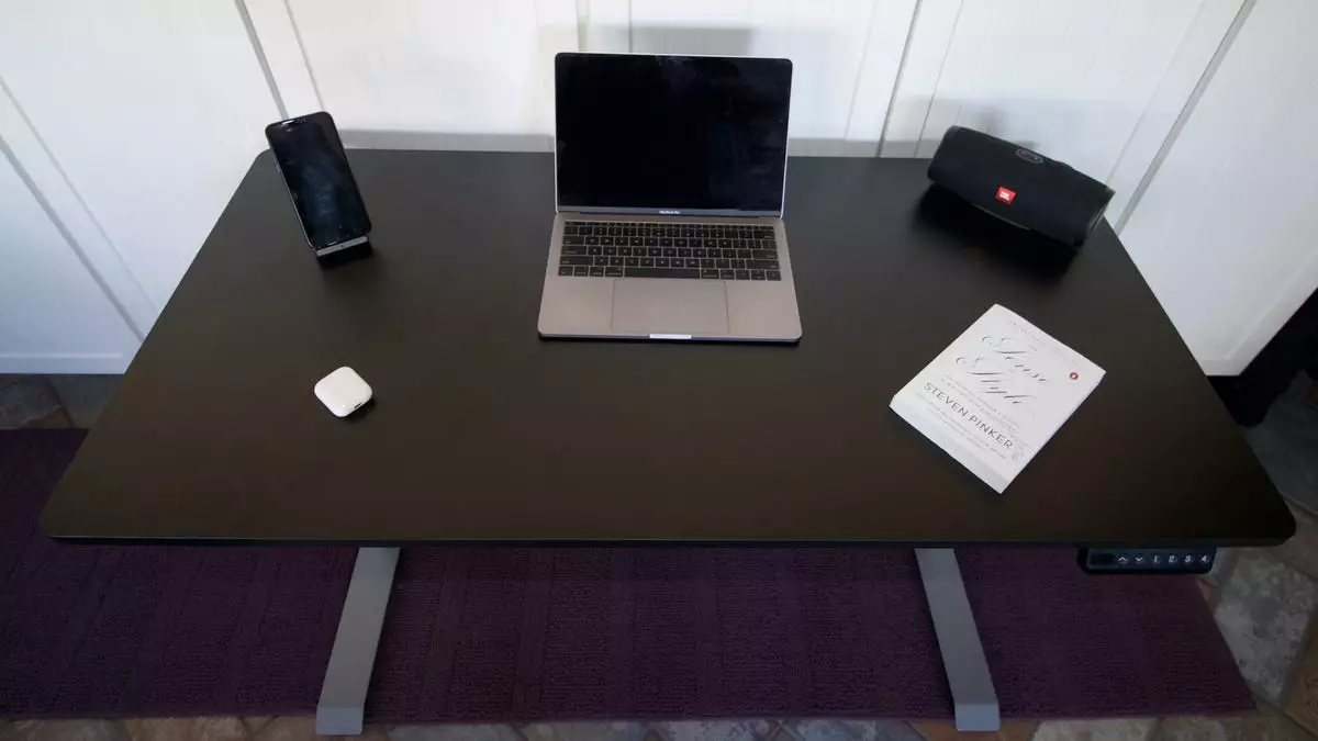 O escritorio de pé Victor High Rise cun portátil, altofalante, teléfono intelixente, AirPods e un libro na superficie.