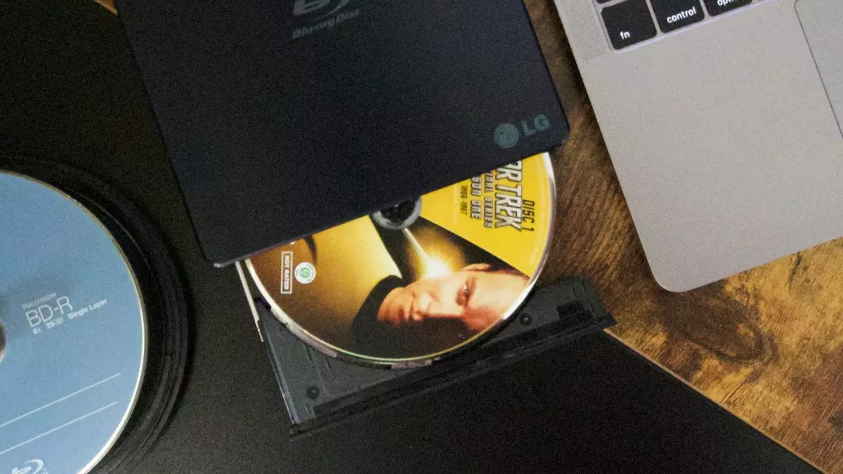 Як шпиндели дискҳои холии Blu-ray, сӯзонандаи диски оптикии беруна ва MacBook Pro дар рӯи миз нишастаанд.