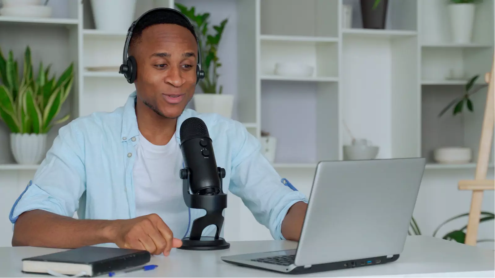 Έννοια Podcast, χαρούμενος νεαρός άνδρας που φοράει ακουστικά και μιλάει σε ένα μικρόφωνο δίπλα στον φορητό υπολογιστή