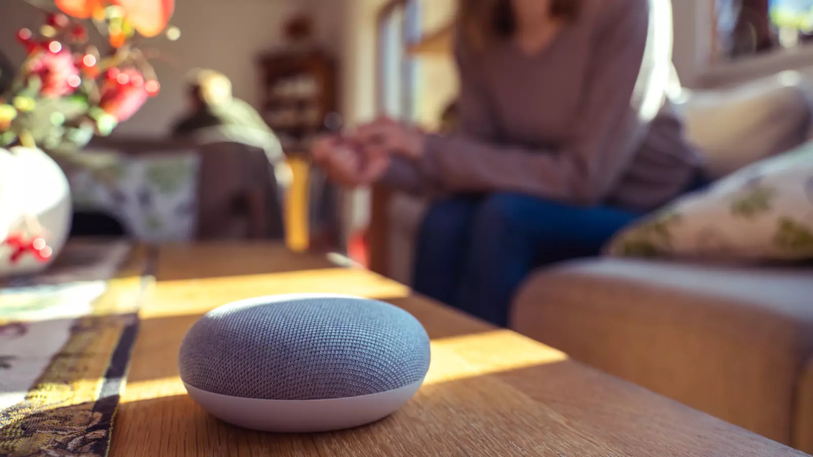 Google Nest Mini viedais skaļrunis uz galda ar cilvēku fonā, kurš sēž uz dīvāna