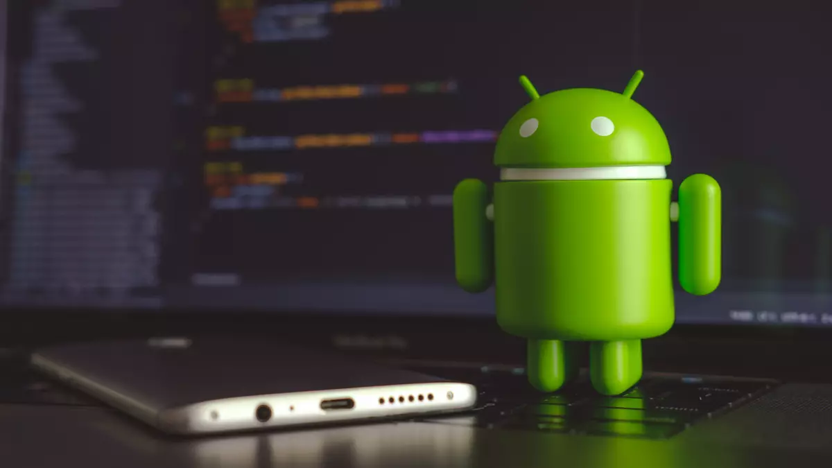 Postava Google Android stojící na klávesnici notebooku vedle telefonu Android s kódem na obrazovce v pozadí