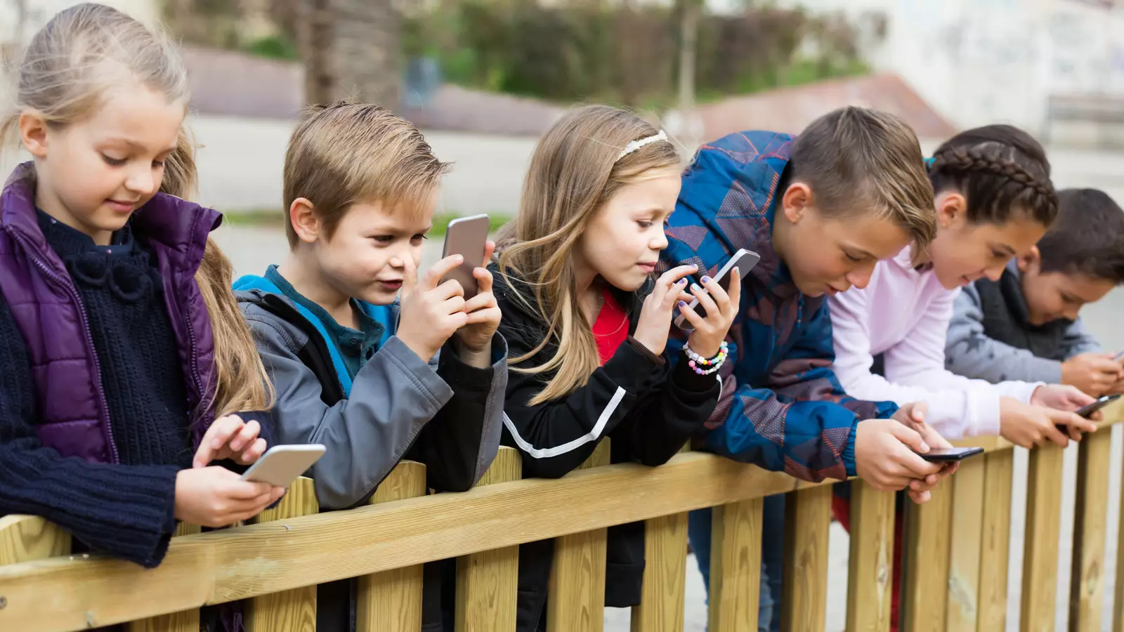 Надворешен портрет на среќни деца кои се потпираат на оградата и гледаат во паметните телефони