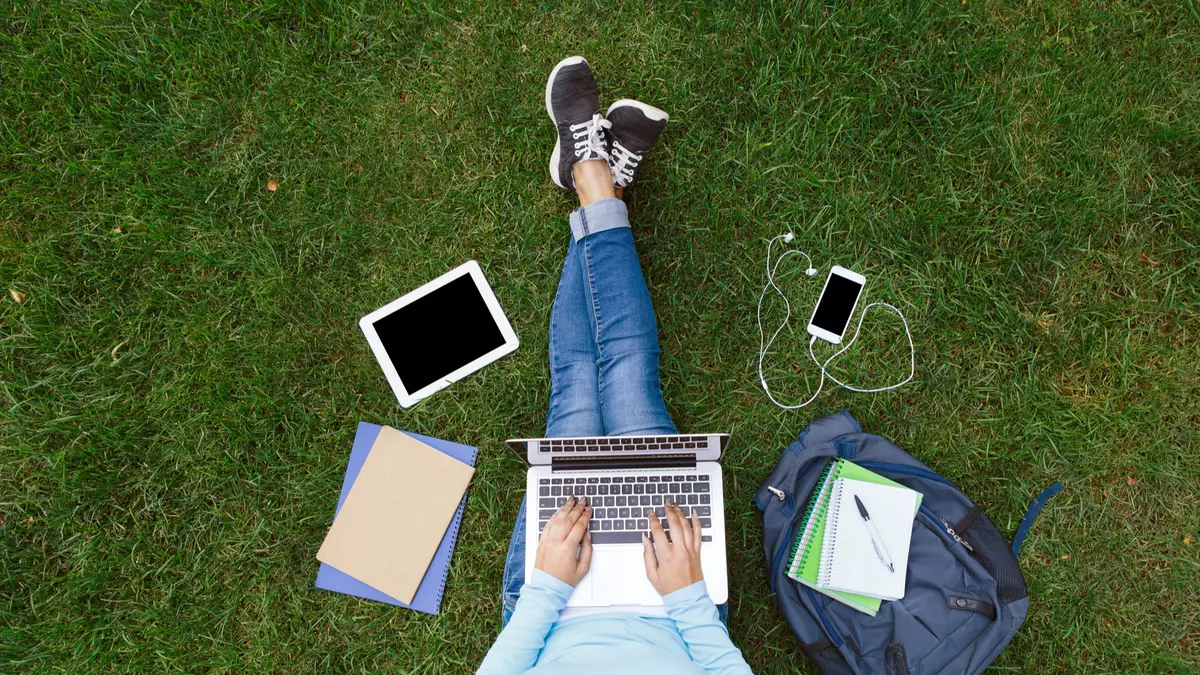 Pohled shora dolů na osobu sedící na trávě v parku s notebookem, tabletem, smartphonem, sluchátky, notebooky a batohem