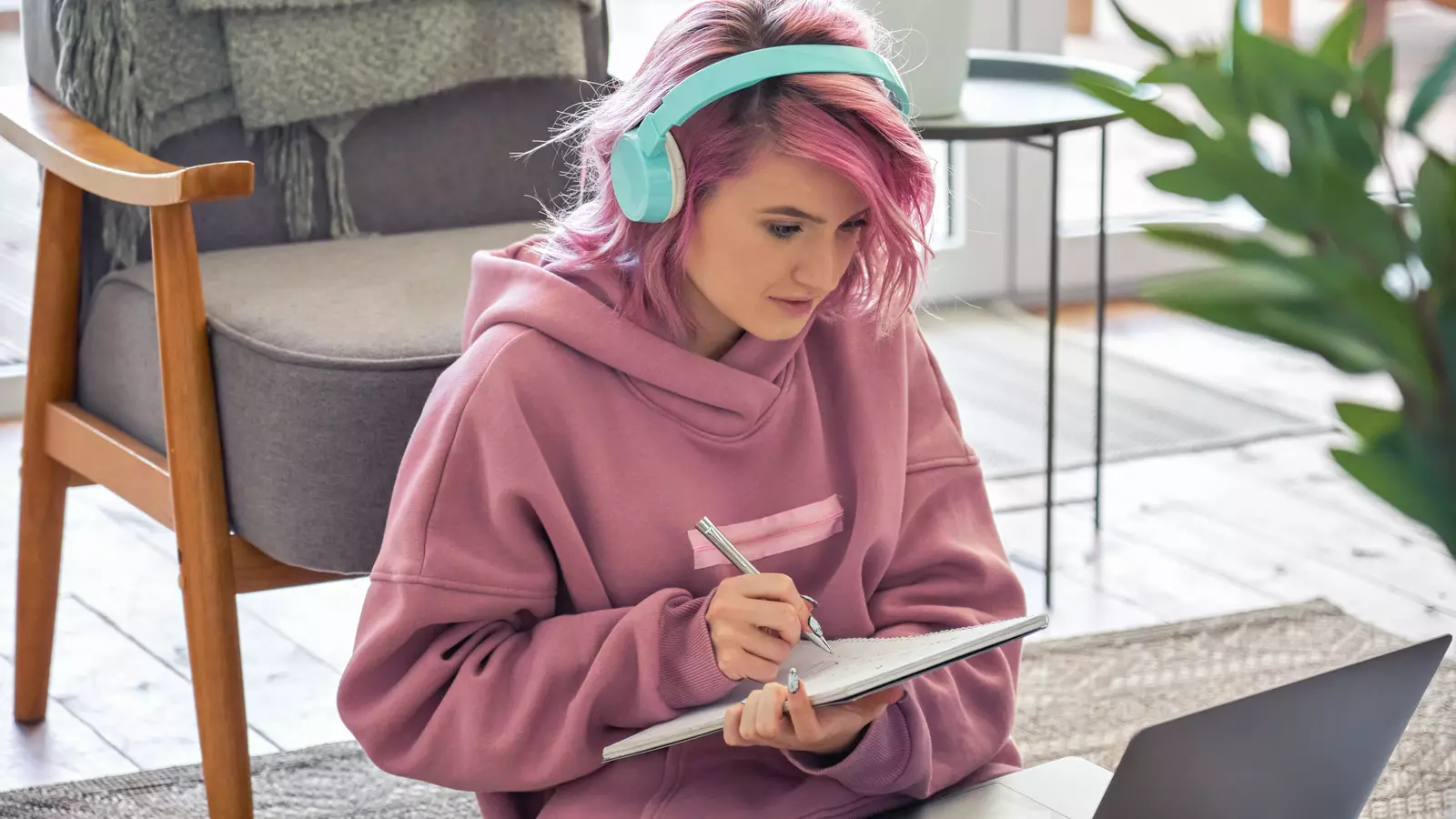 Student van middelbare leeftijd met roze haar die een koptelefoon draagt en aantekeningen maakt tijdens het kijken naar een video op een laptop