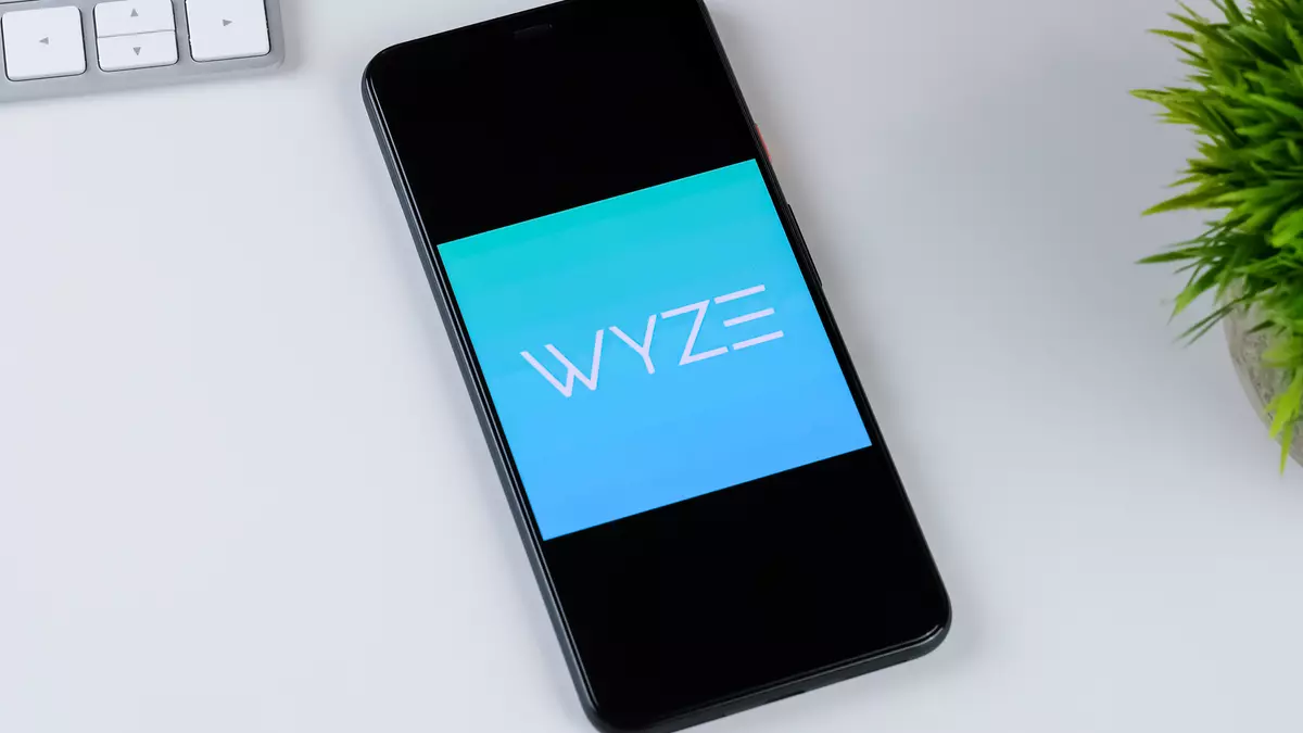Wyze app-logotyp på en smartphoneskärm som ligger på ett skrivbord med ett tangentbord och en växt