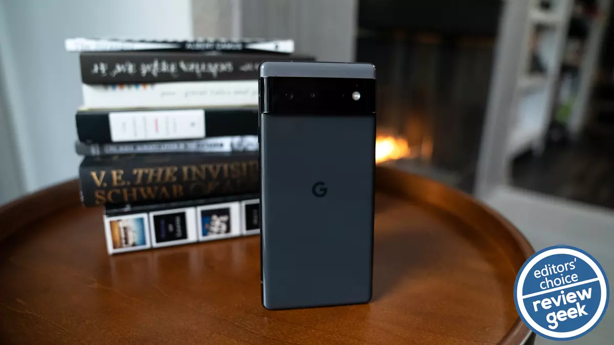 কাঠের টেবিলে Google Pixel 6 স্মার্টফোনটি ব্যাকগ্রাউন্ডে ফায়ারপ্লেস সহ বইয়ের স্তুপের সাথে ঝুঁকে আছে