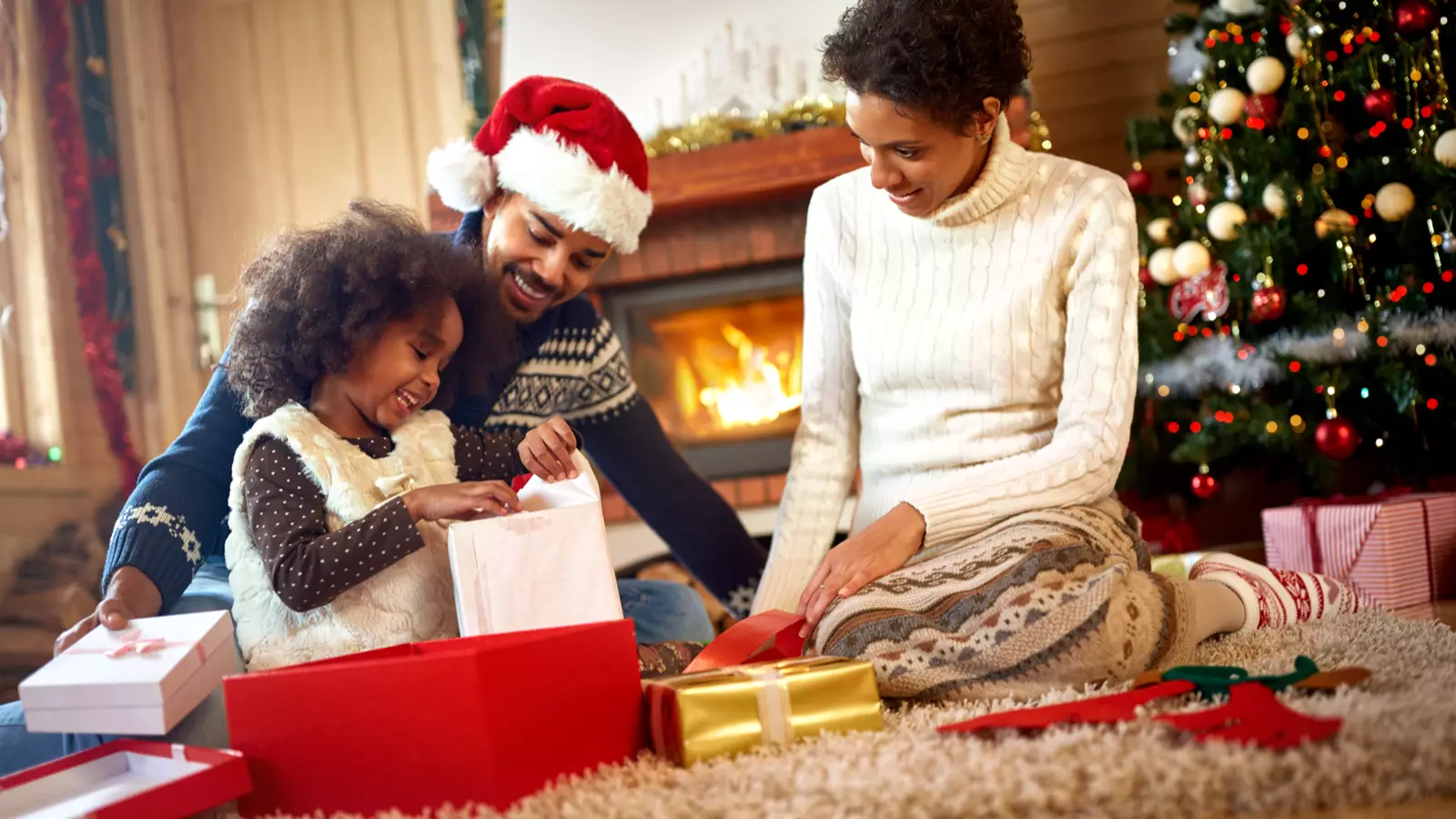 Cha mẹ nhìn con mình mở quà Giáng sinh trước lò sưởi và cây thông Noel