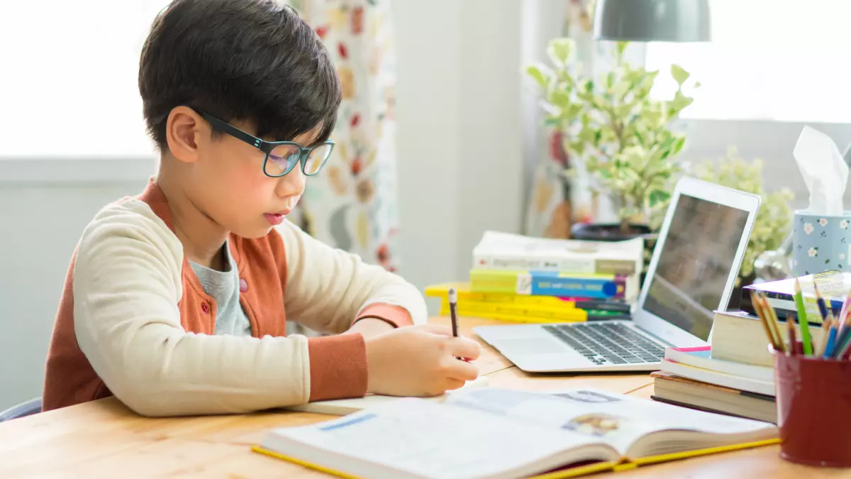 En smart preteen-dreng sidder ved et bord og skriver lektier og forbereder sig til en online eksamen