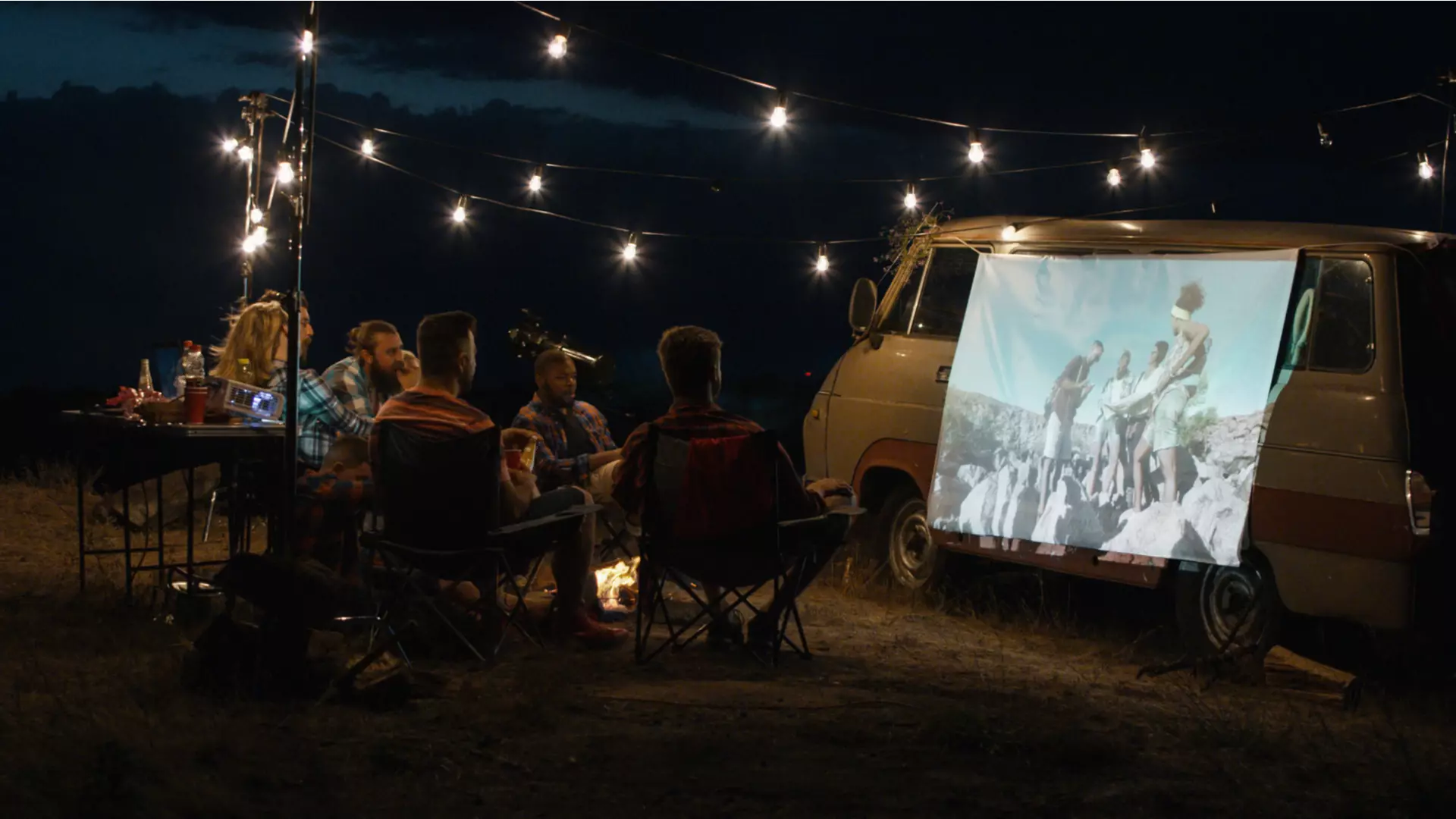 Prijatelji se zbirajo v kampu okoli kresa in gledajo film s projektorjem na strani kombija v temnem večeru