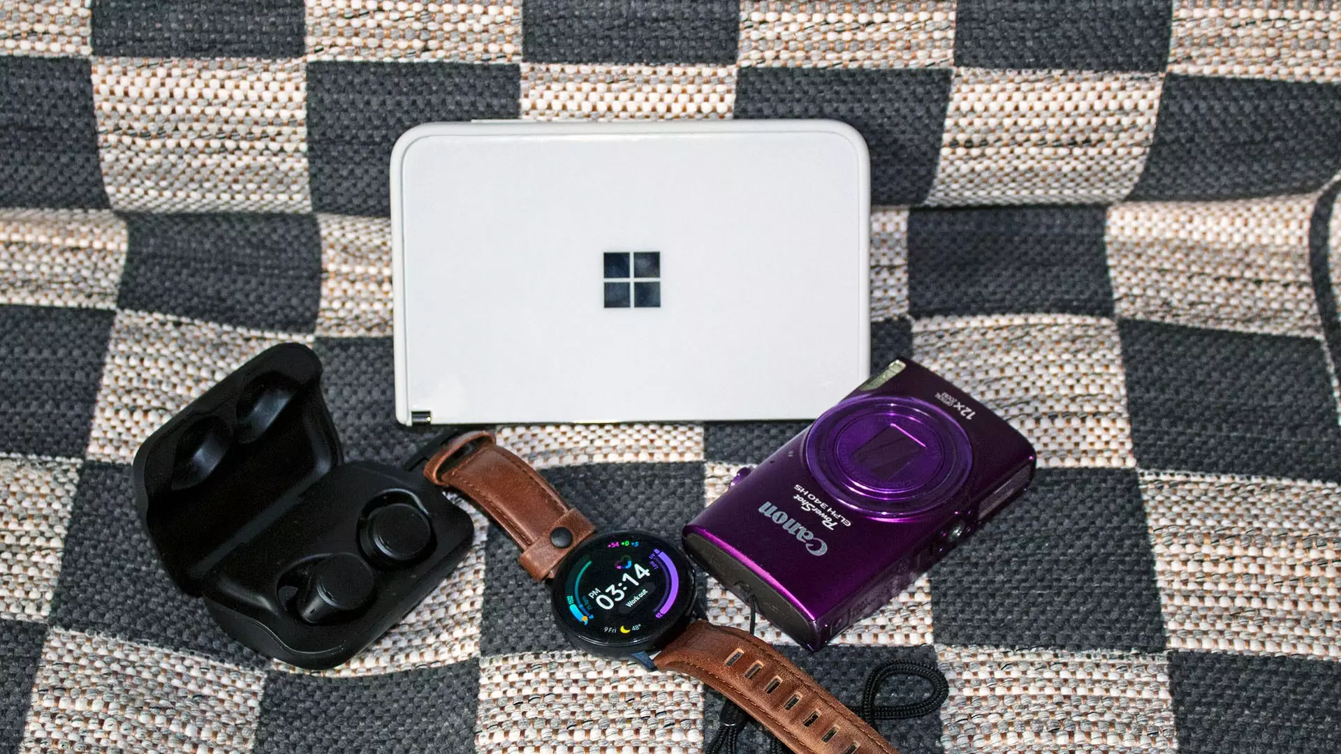 Surface Duo, pametni sat, ručna kamera i prave bežične slušalice na kariranoj dekici.