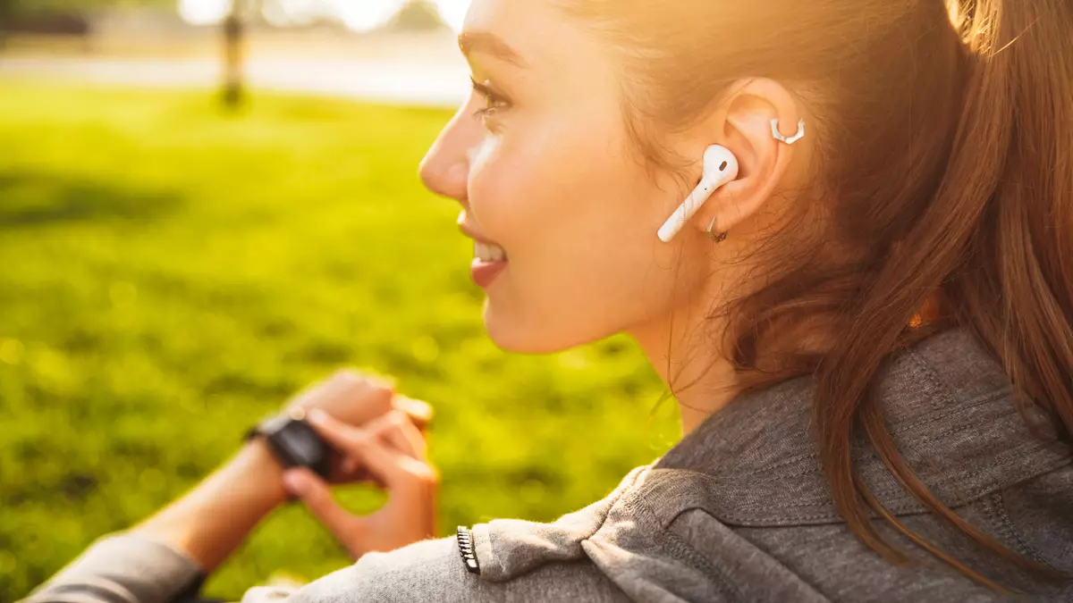 Wanita sporty berusia 20-an dalam pakaian sukan menggunakan jam tangan pintar dan fon telinga wayarles semasa berehat di taman