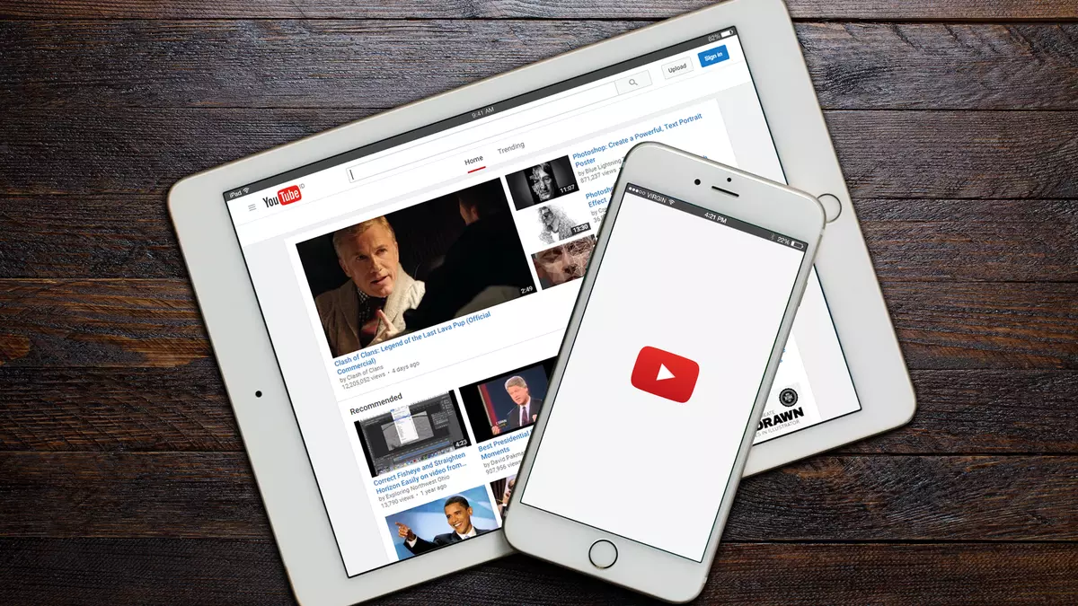 Skrin memuatkan apl YouTube pada iPhone dan halaman pendaratan tapak web YouTube pada iPad kedua-duanya terletak di atas meja kayu