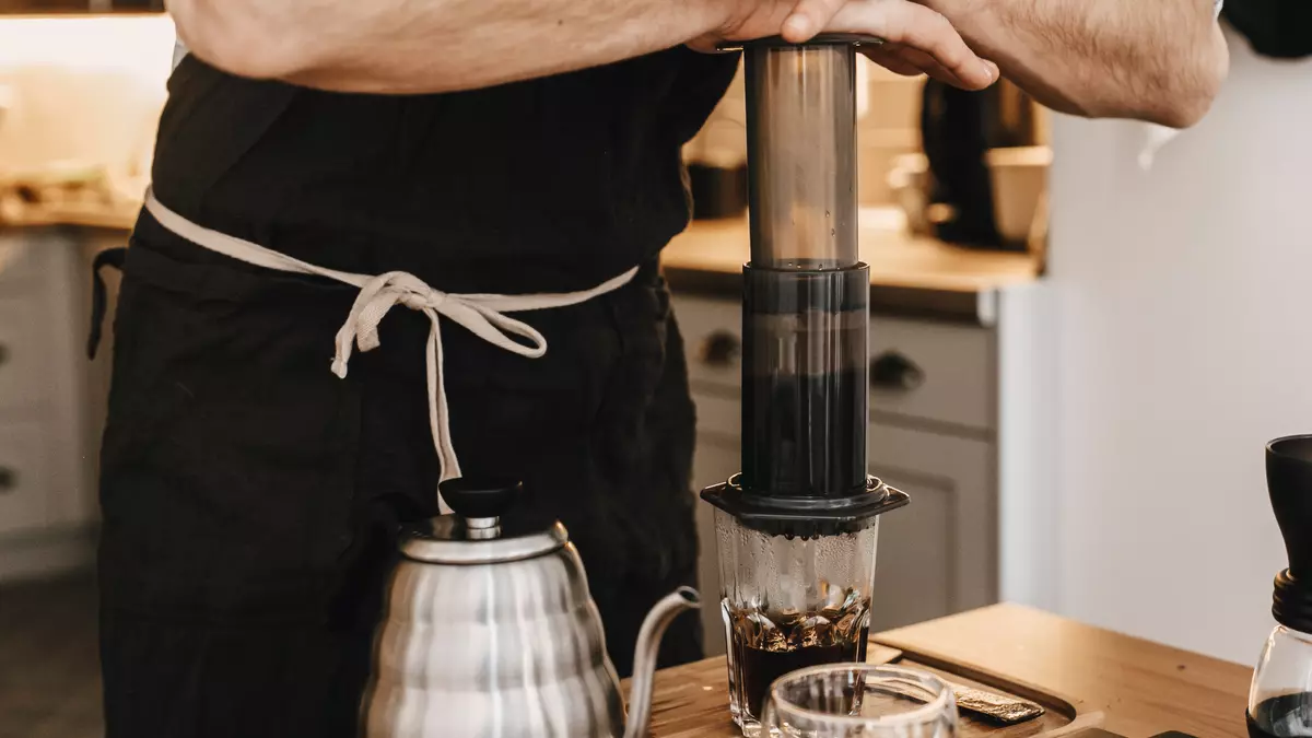 Profesionální barista připravující kávu v aeropressu, alternativní způsob vaření kávy. Ruce na aeropress a skleněný pohár, váhy, ruční mlýnek, kávová zrna, konvice na dřevěný stůl