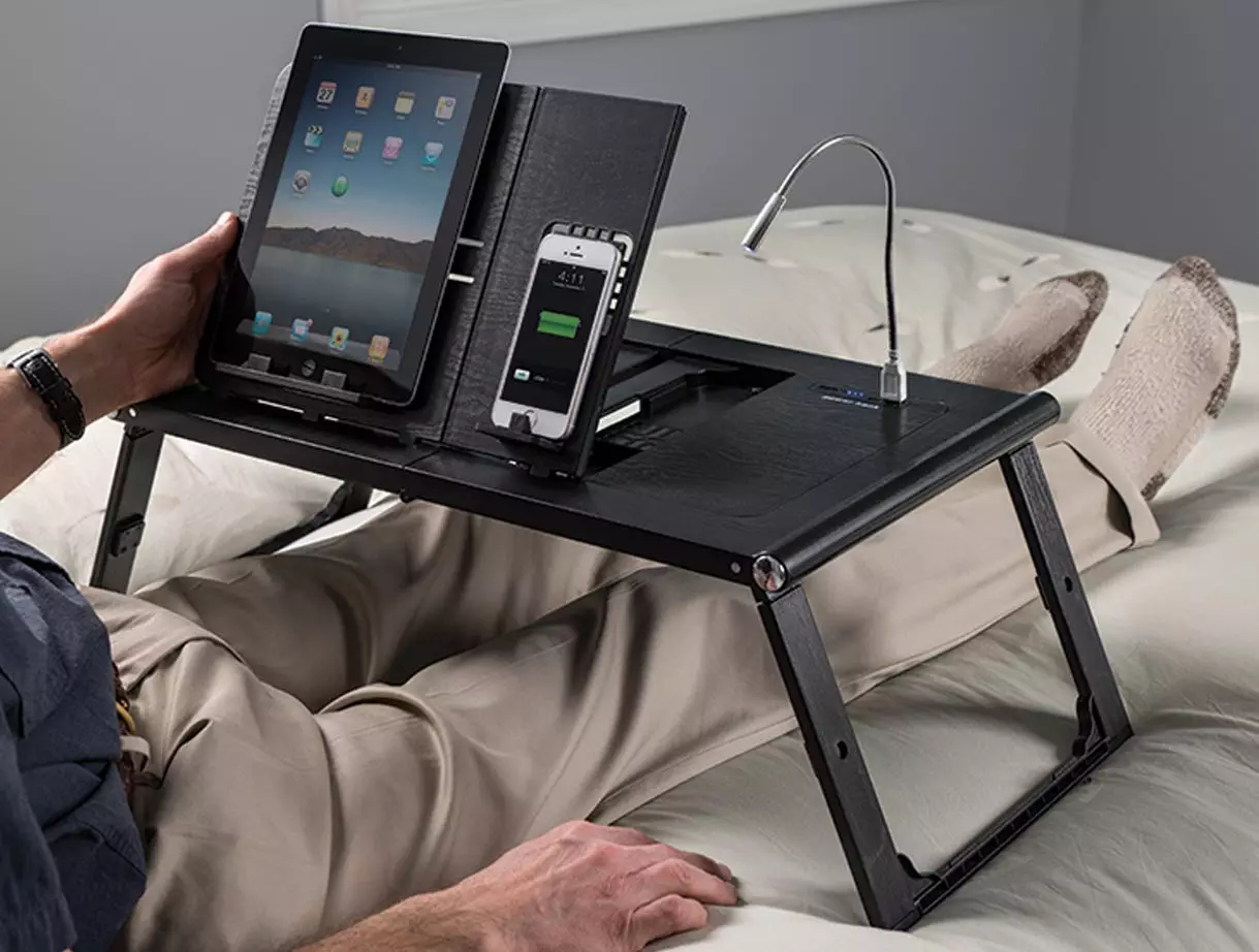 Il vassoio per laptop e tablet Sharper Image su un letto, con le gambe distese di un uomo sotto, la mano appoggiata su un tablet sul vassoio.