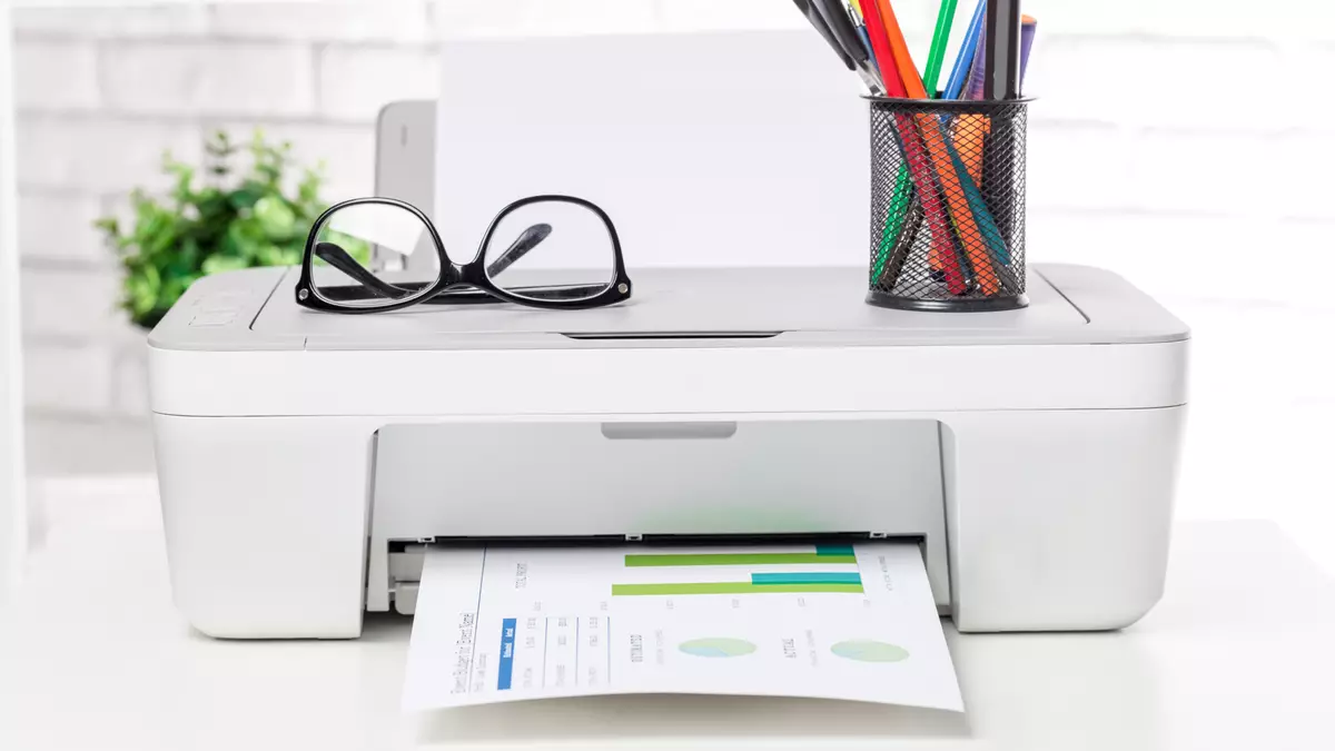 Një printer mbi një tavolinë me letra, syze dhe enë shkrimi