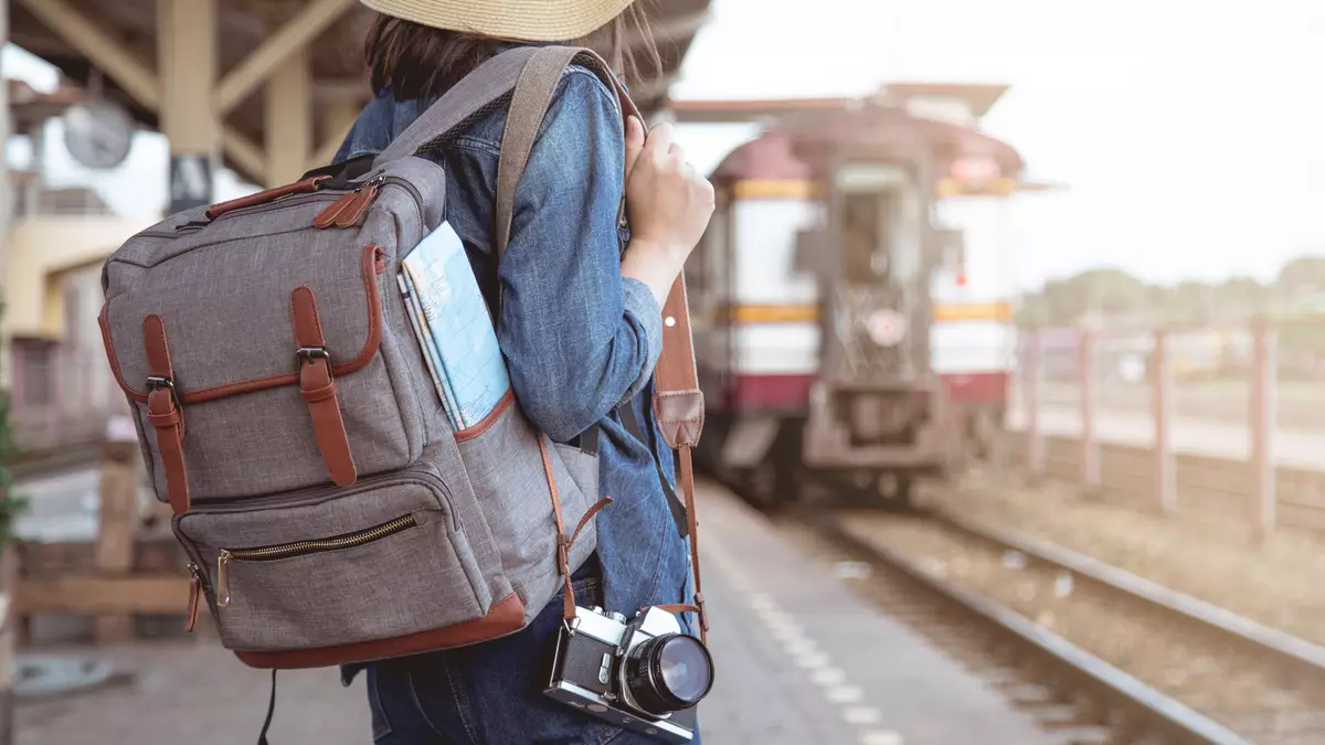 Kobieta z torbą i aparatem fotograficznym przewieszonym przez ramię, czekająca na wjazd pociągu na stację.