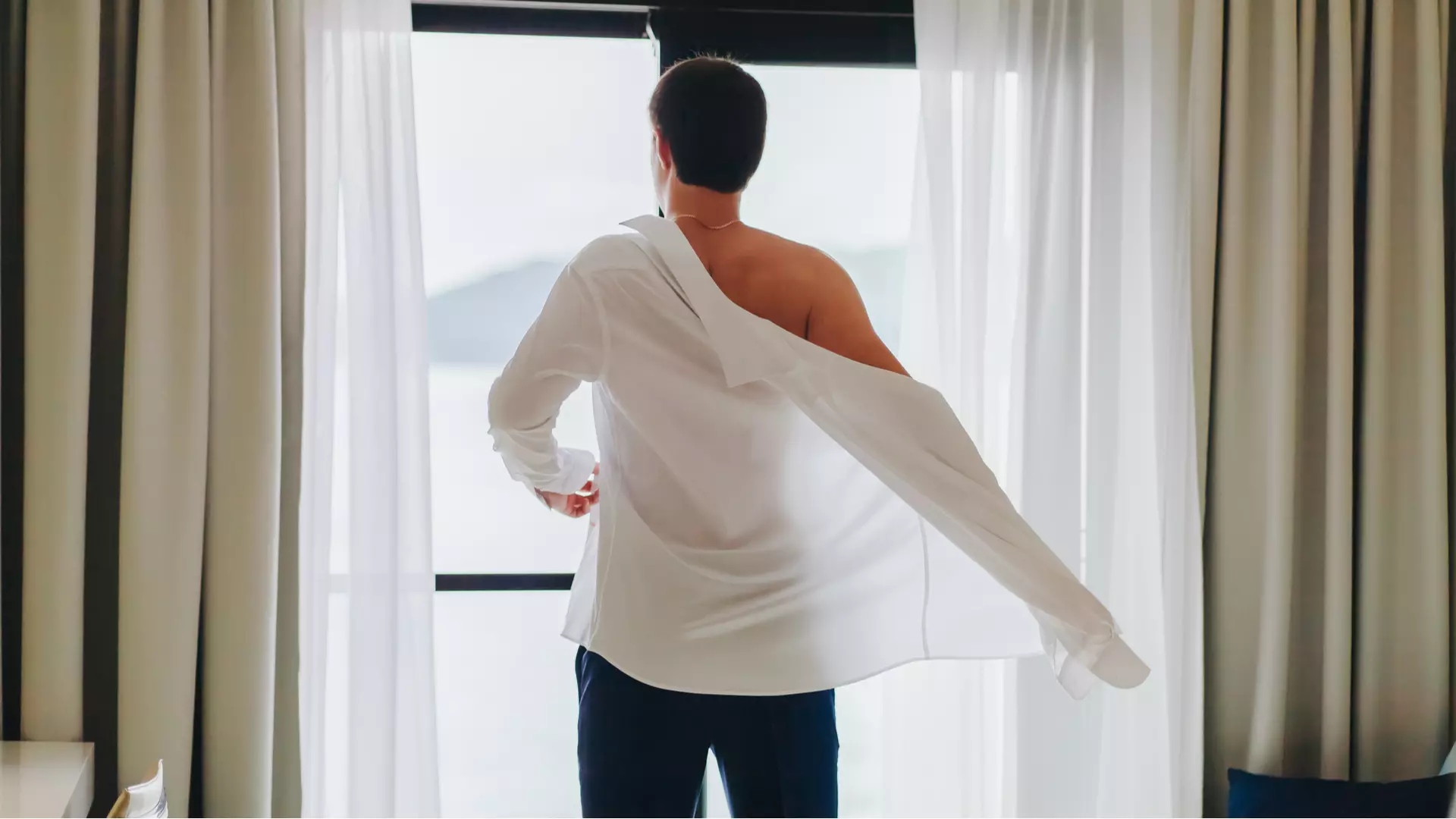 Muž si pred oknom oblieka košeľu s gombíkmi