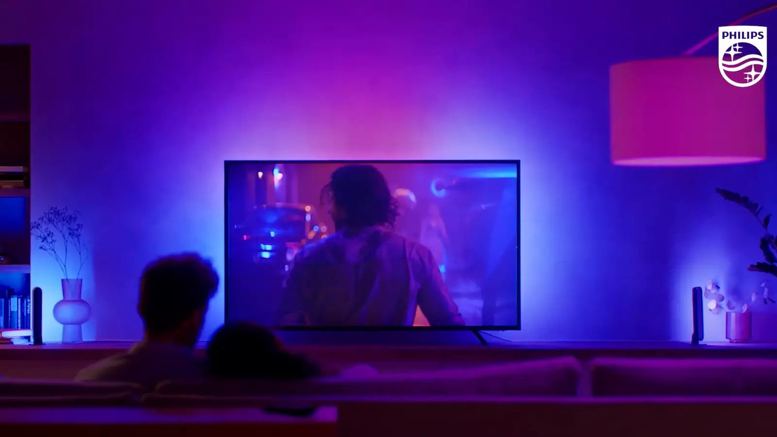 Philips Hue Play Gradient Lightstrip, რომელიც ასახავს ფერებს ტელევიზორის უკან კედელზე, წინა პლანზე დივანზე მჯდომი წყვილი და უყურებს მას