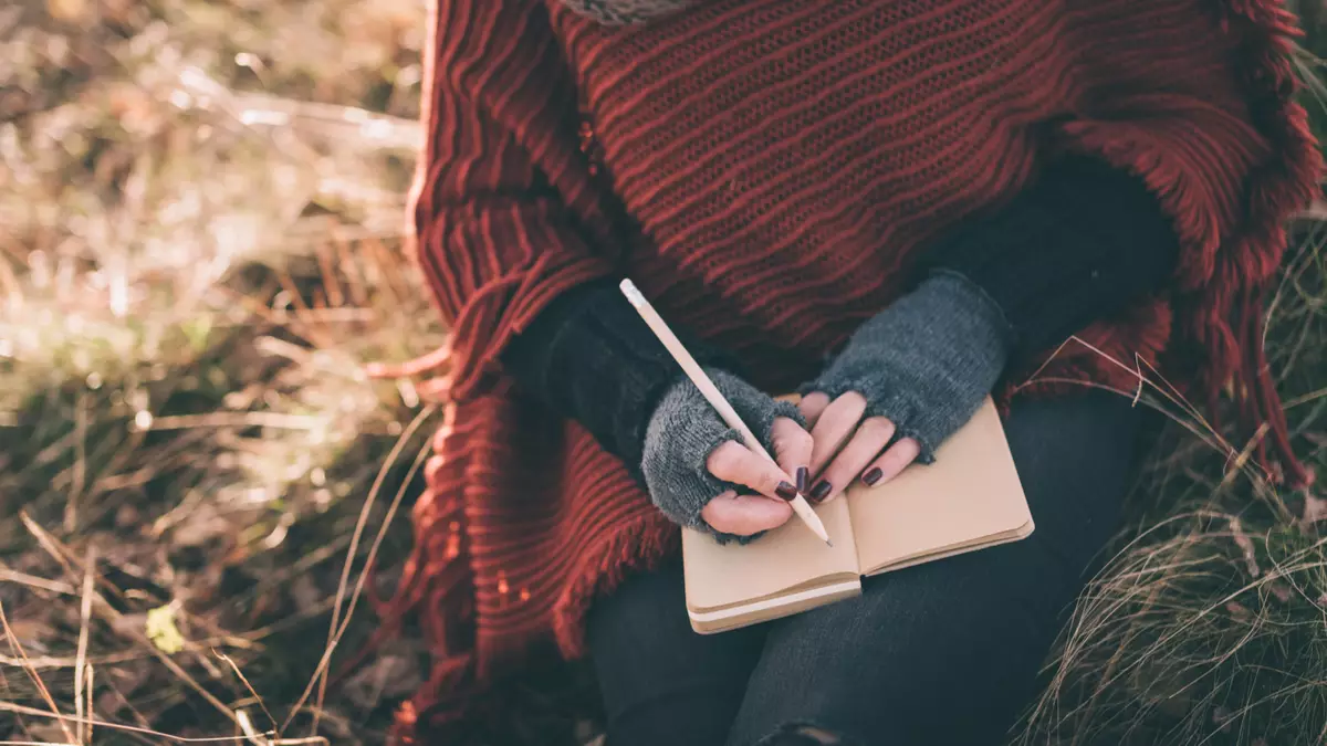 Een vrouw met een potlood boven een opengeslagen notitieboekje op haar schoot.