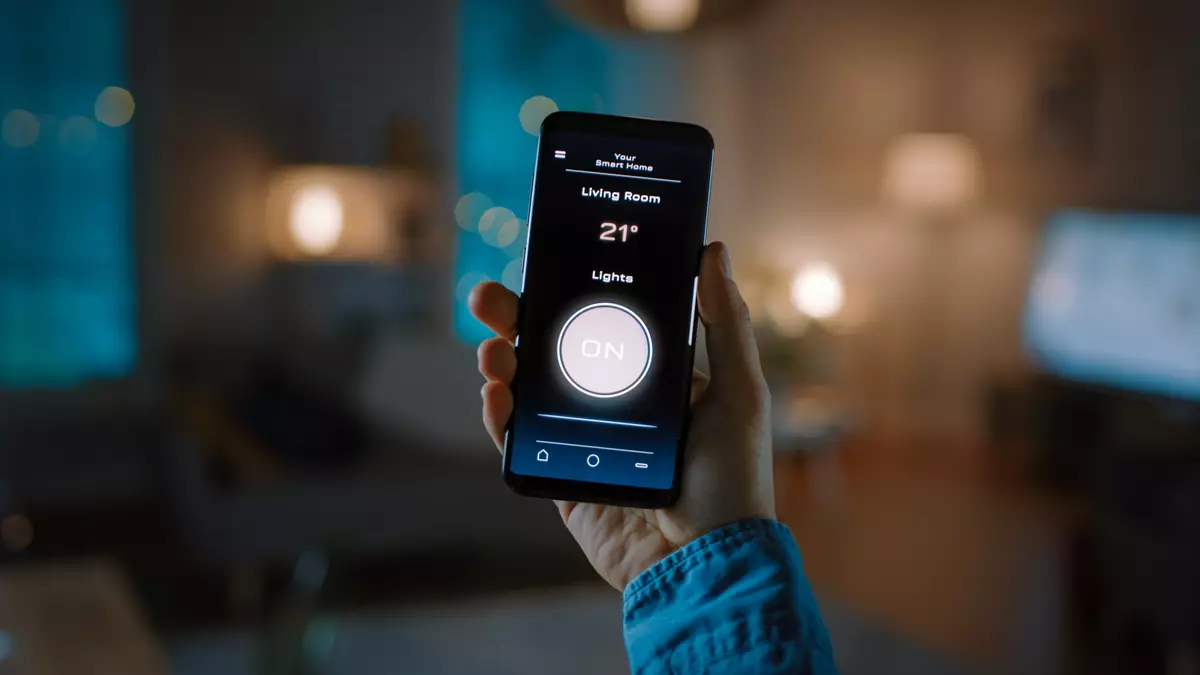सक्रिय स्मार्ट होम ऍप्लिकेशनसह स्मार्टफोनचा क्लोज अप शॉट. खोलीतील दिवे चालू/बंद करण्यासाठी व्यक्ती व्हॉइस कमांड देत आहे. अपार्टमेंटमध्ये आरामदायक संध्याकाळ आहे.