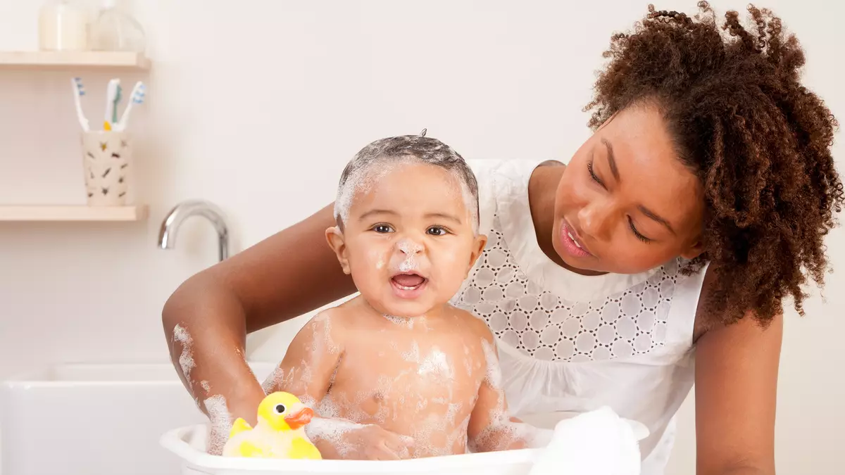 En baby täckt av tvål som sitter i ett litet badkar medan hans mamma badar honom.
