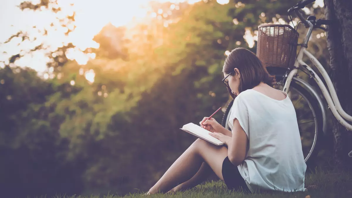 Жінка сидить у траві, пише в журналі