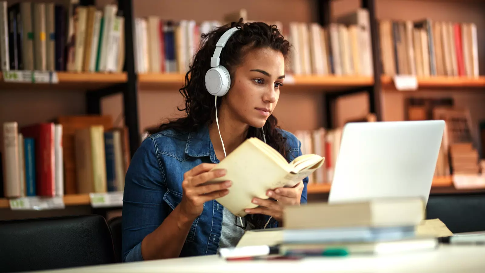 Млада жена у библиотеци држи књигу и гледа у свој лаптоп.