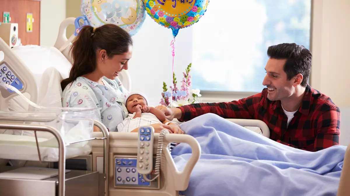 Unha muller nunha cama de hospital sostendo a un recén nacido mentres o pai lle toca amorosamente o brazo pequeno