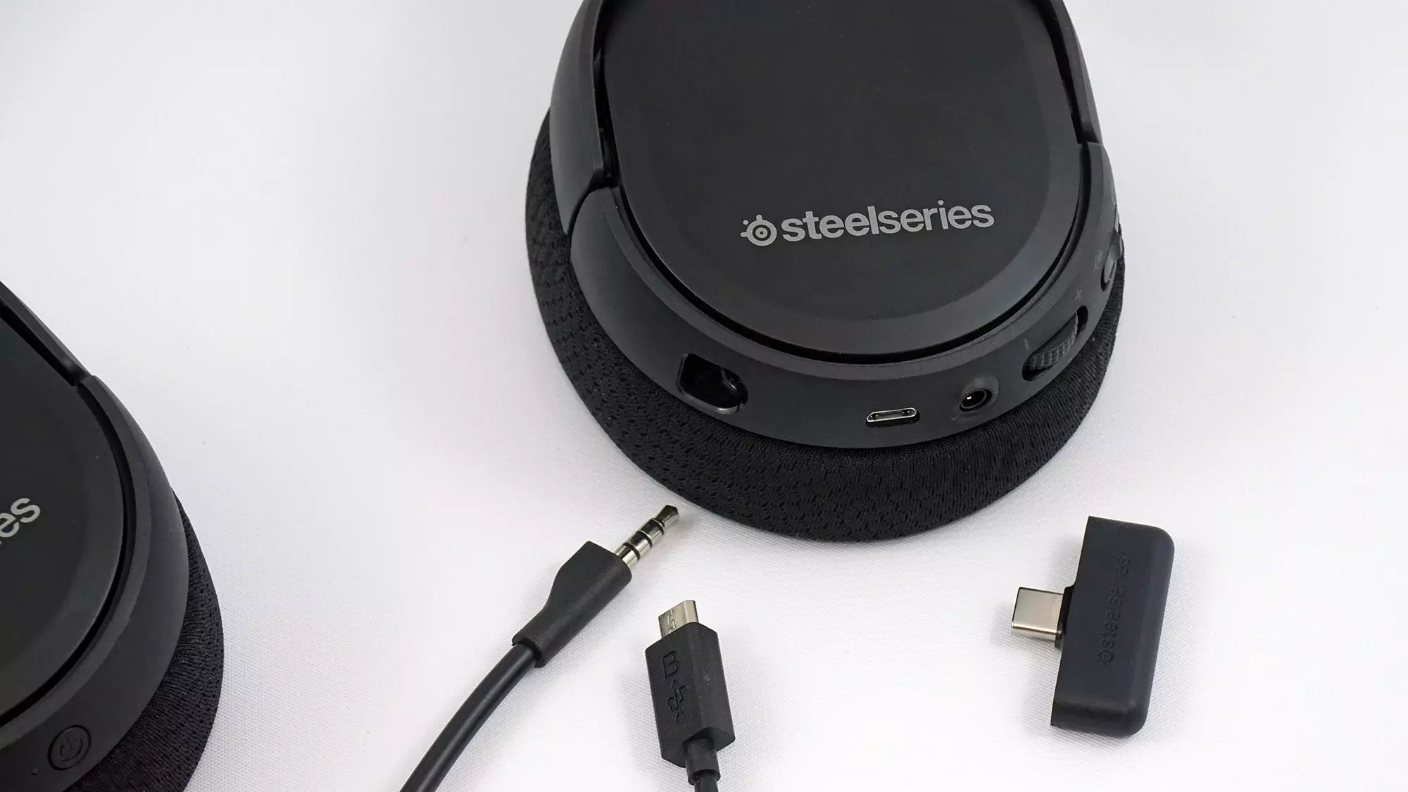 Arctis 1 Kablosuz kulaklıklardan birinin yanında bir USB-C dongle ve MicroUSB şarj kablosu.
