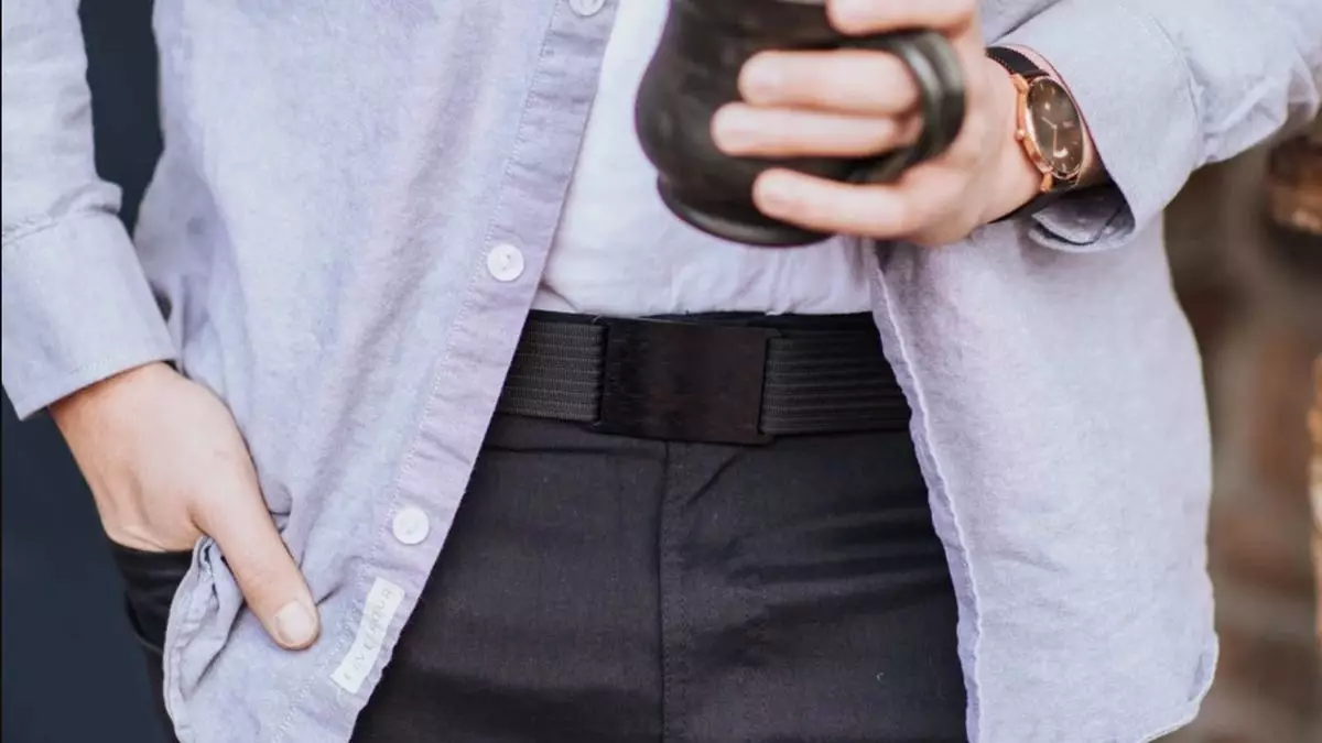 Bir kahve kupası tutan ve koyu renk pantolonun üzerine siyah bir Grip6 kemeri takan bir adamın eli.