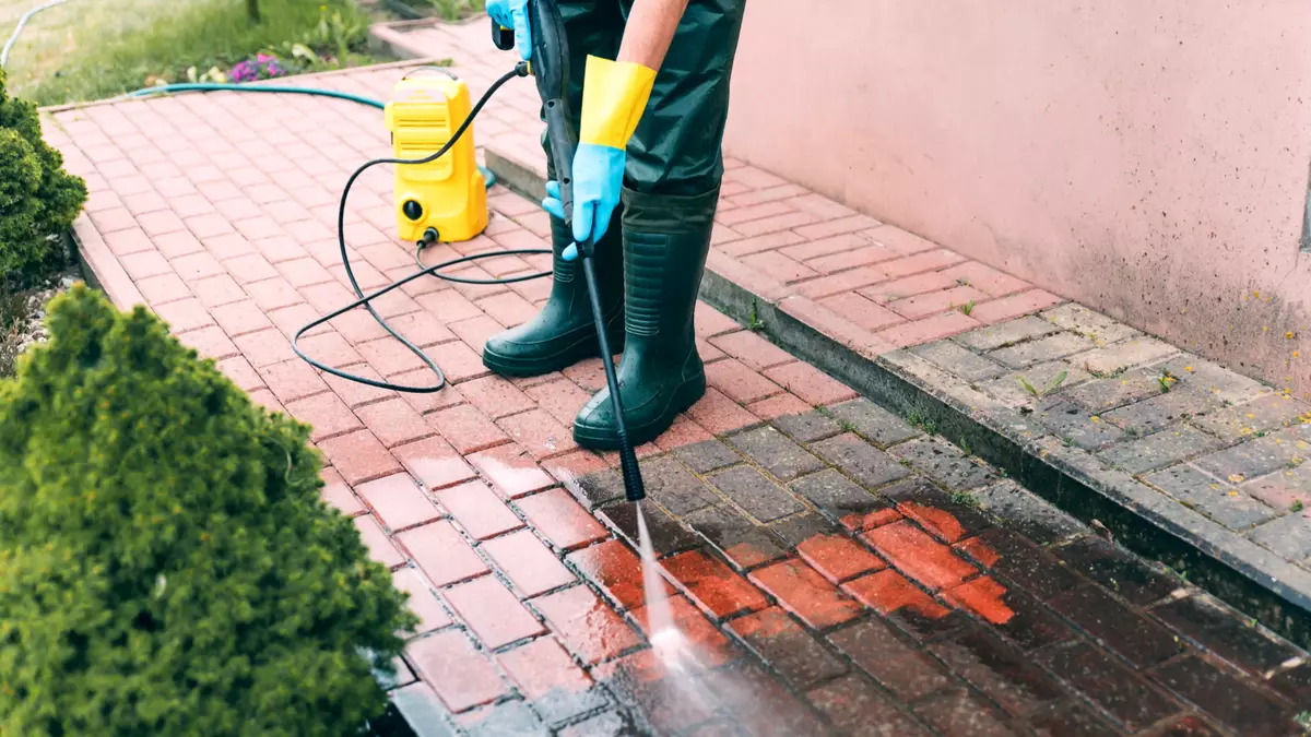 مردی در حال تمیز کردن بلوک پیاده رو قرمز و بتنی با استفاده از آب پاک کن فشار قوی. مفهوم تمیز کردن سنگفرش. مردی با پوشیدن شلوار، شلوار محافظ، ضد آب و دستکش در حال انجام کارهای بهاری در باغ.