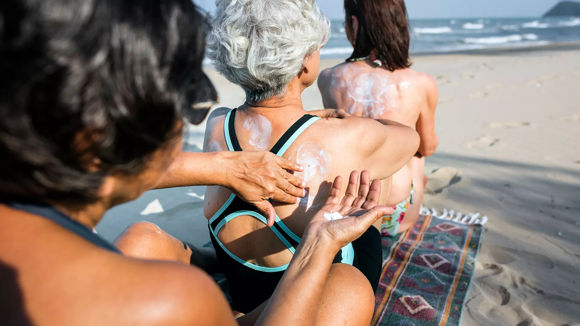Tři ženy na pláži si navzájem nanášejí opalovací krém na záda.