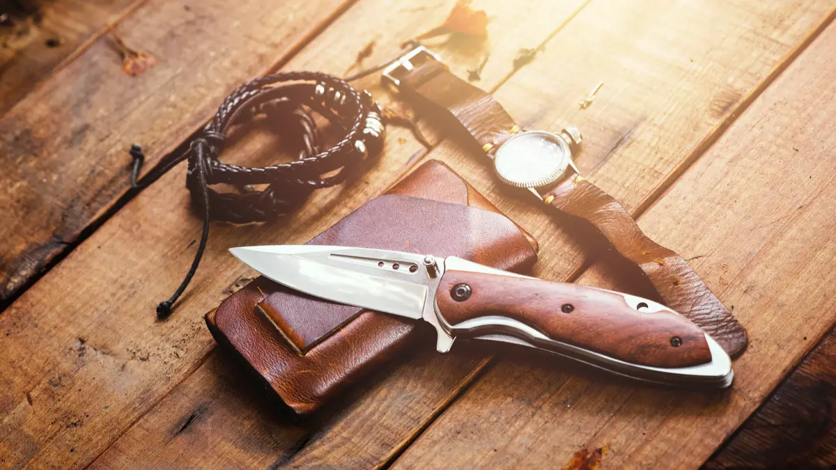 Un cuchillo premium sobre una cartera, junto a un reloj y una pulsera de cuero sobre una mesa de madera.