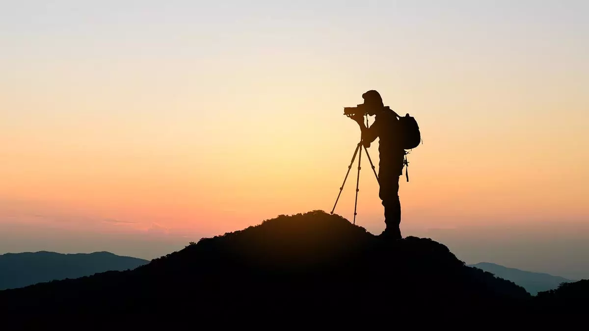 प्रवास फोटोग्राफी गियरसह पर्वताच्या शिखरावर उभी असलेली व्यक्ती, लँडस्केप छायाचित्र घेत आहे